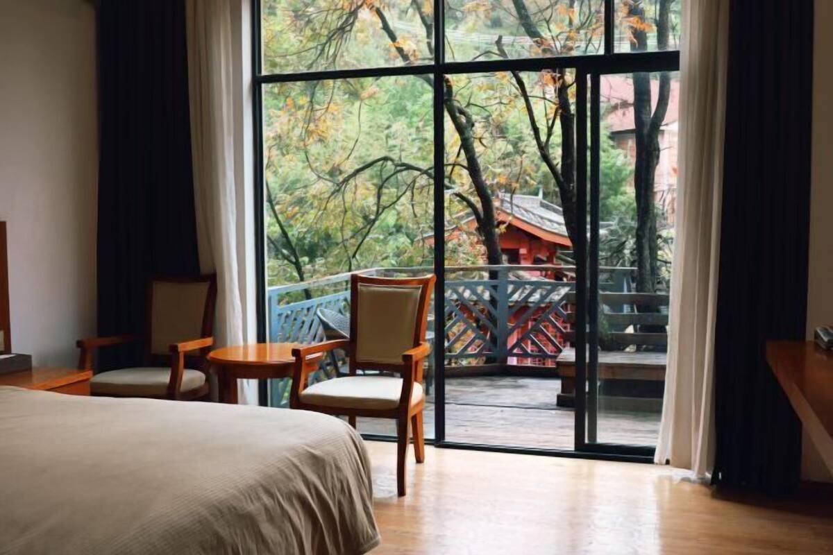 Quarto de hotel com cama, duas cadeiras, uma mesa pequena e uma grande porta de vidro deslizante que dá para área externa com vista para árvores e um edifício tradicional.