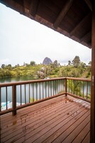 Um deck de madeira com guarda-corpo de metal, com vista para um lago tranquilo, rodeado por vegetação e uma montanha imponente ao longe.