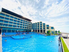 Uma ampla área de piscina ao ar livre, com várias piscinas e um edifício moderno com grandes janelas ao fundo.