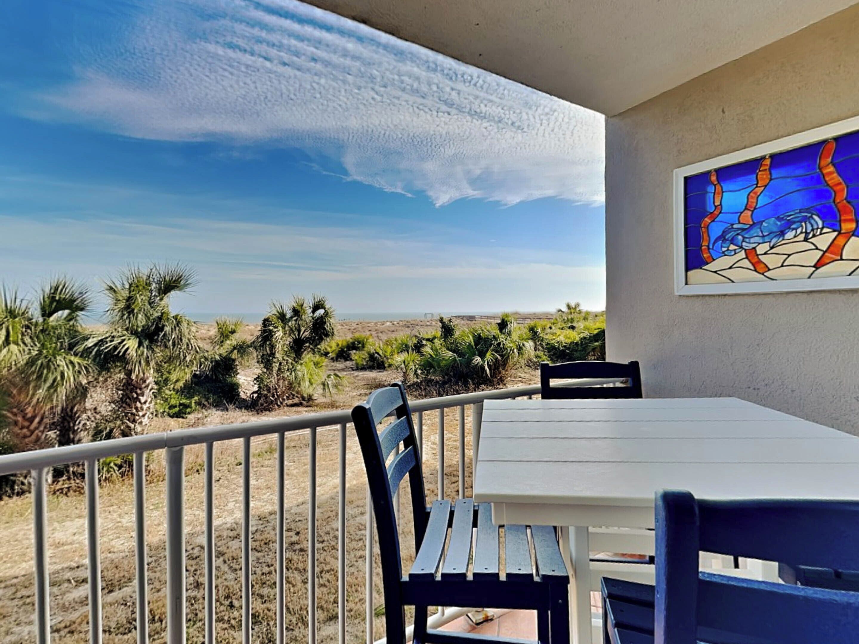 Varanda com mesa e cadeiras brancas, vista para uma praia com palmeiras e céu claro.