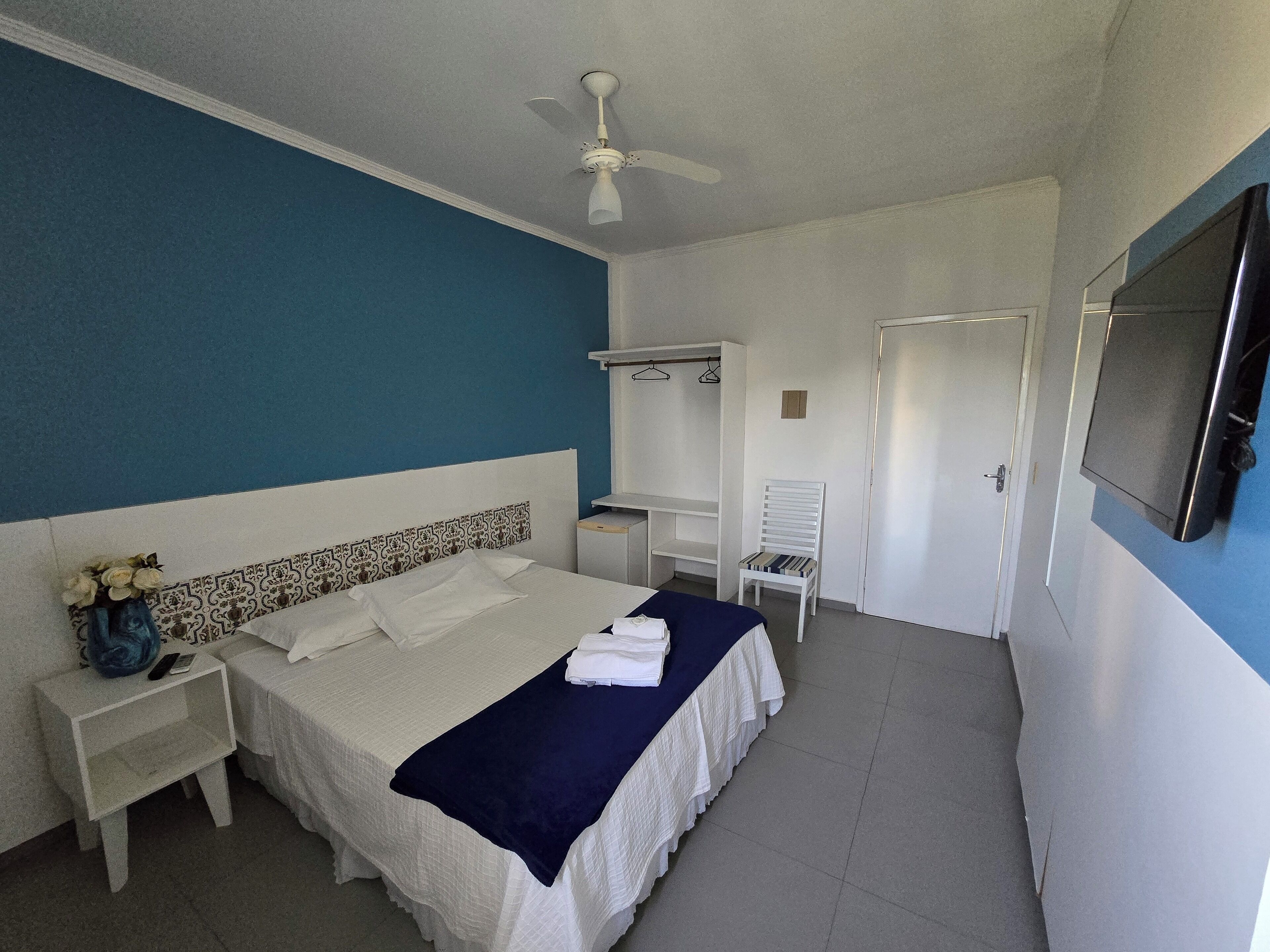 Um quarto com uma cama, mesa de cabeceira, cadeira, guarda-roupa e televisão.
