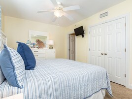 Quarto com cama, ventilador de teto, televisão na parede e uma porta.