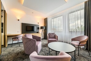 Quarto de hotel moderno com televisão de tela plana, escrivaninha e área de estar com cadeiras acolchoadas e uma mesa redonda.