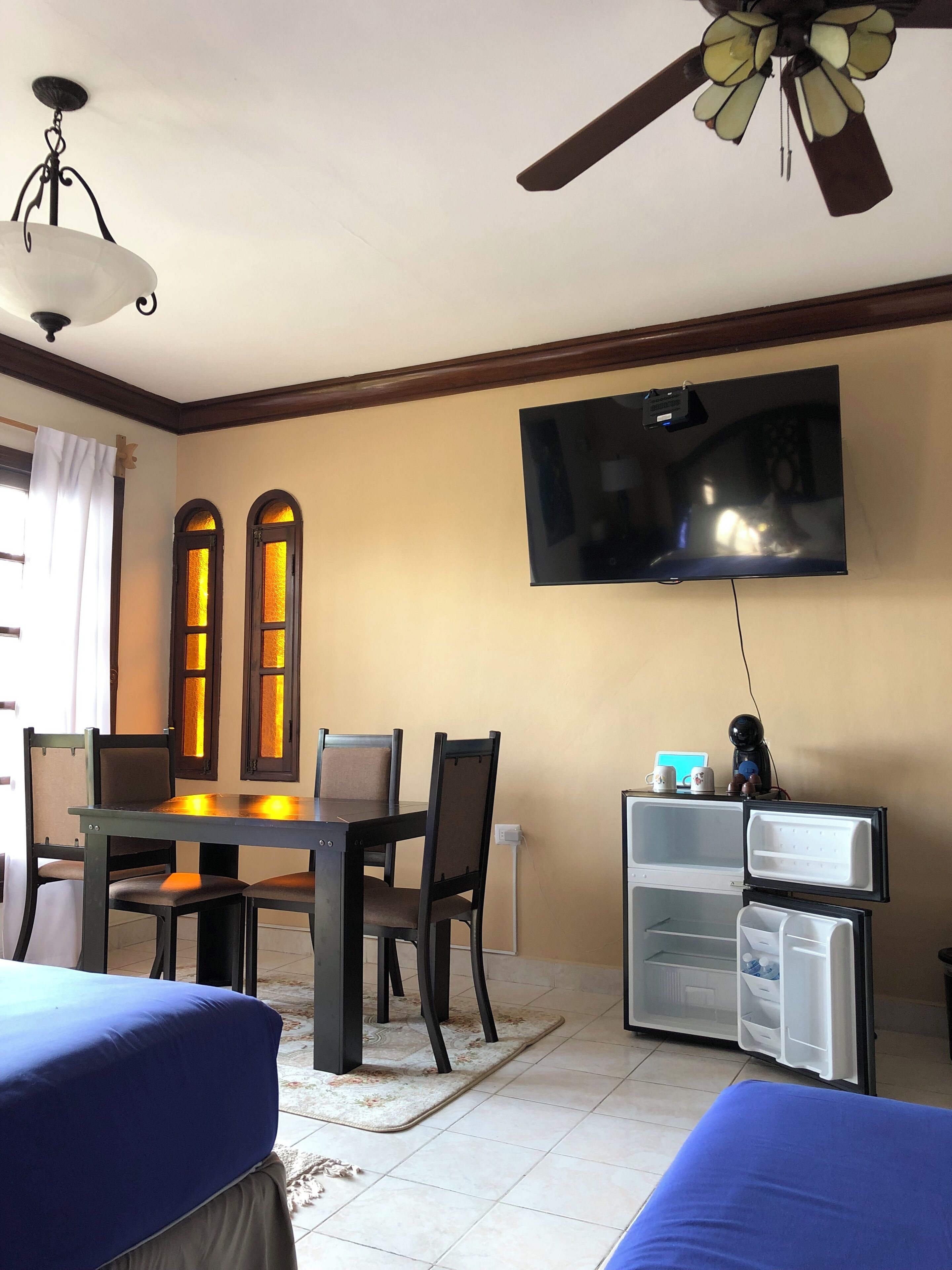 Quarto com mesa de jantar, cadeiras, ventilador de teto, TV na parede e mini-geladeira.