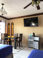 Quarto com mesa de jantar, cadeiras, ventilador de teto, TV na parede e mini-geladeira.