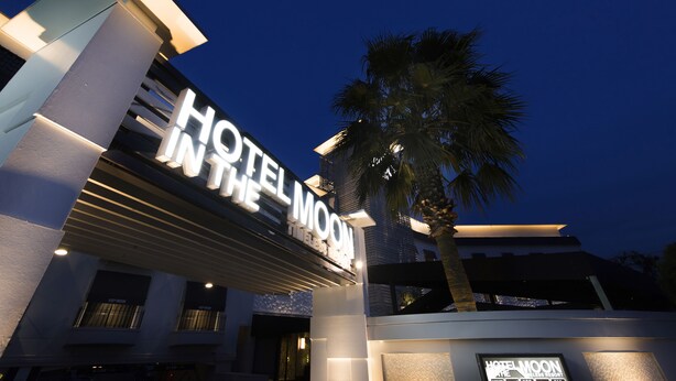 Exterior do HOTEL MOON, com uma placa destacada, arquitetura moderna e uma palmeira.