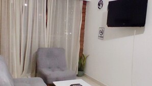 Uma sala de estar moderna com duas poltronas cinzas, uma mesinha branca e uma televisão de tela plana na parede.