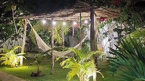 Área externa bem iluminada com uma rede, palmeiras e uma pérgola com luzes de cordão.
