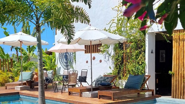 Piscina com espreguiçadeiras e guarda-sóis, rodeada por vegetação e um edifício branco com um deck de madeira.