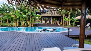 Um deck de madeira com piscina, espreguiçadeiras e uma estrutura coberta de palha.