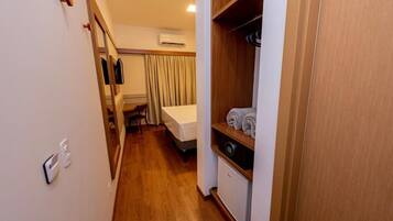 Corredor estreito que leva a um quarto com cama, uma mesinha e uma cadeira. Há um guarda-roupa de madeira com uma prateleira contendo toalhas. Um aparelho de ar condicionado está fixado na parede acima da cama.