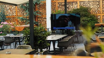 Área externa com mesas e cadeiras, uma tela grande exibindo uma programação, e uma parede decorada com troncos de madeira e vegetação.