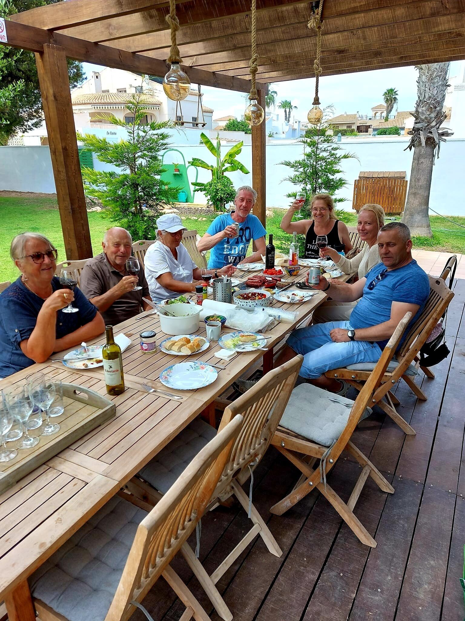 Um grupo de pessoas jantando em uma mesa de madeira ao ar livre, com vista para a água e edifícios ao fundo.