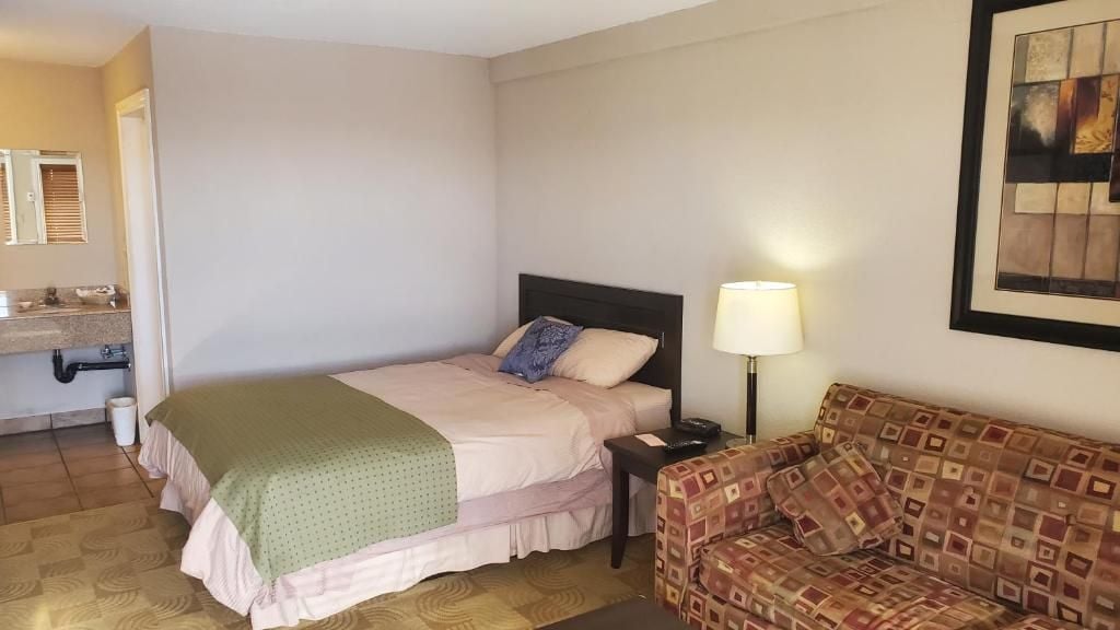 Quarto de hotel com cama, mesa de cabeceira, luminária, sofá e banheiro ao fundo.