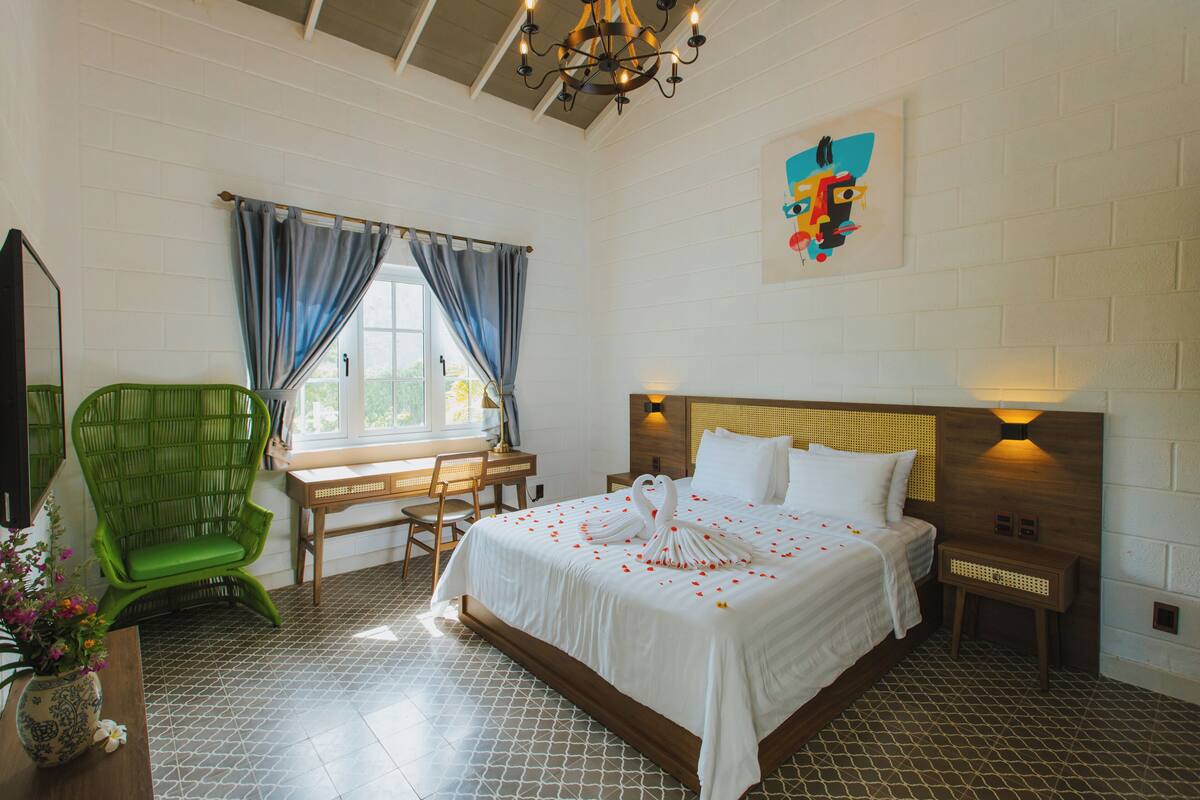 Quarto de hotel com uma cama grande, uma cadeira verde, uma escrivaninha e uma janela com cortinas.