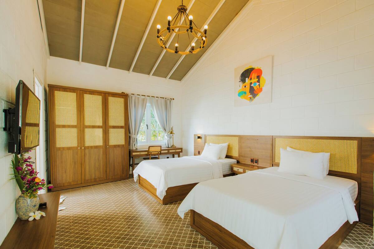 Quarto de hotel com duas camas, teto de madeira, lustre, escrivaninha com cadeira, janela com cortinas e televisão na parede.