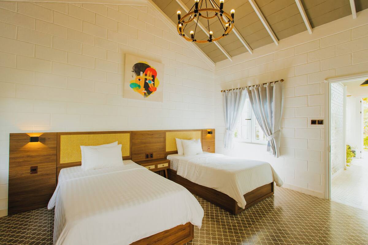 Quarto de hotel com duas camas, um candelabro, um quadro e vista para o exterior.