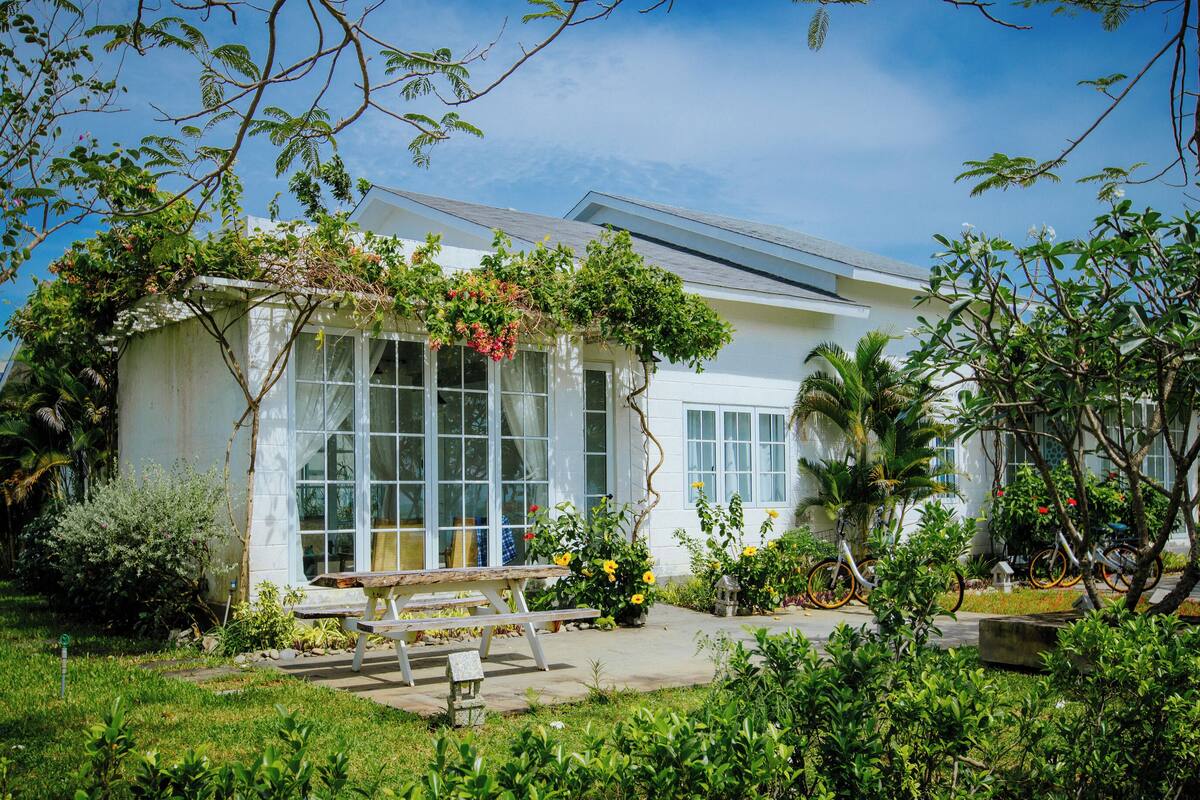 Uma casa branca com grandes janelas, um alpendre com mesa e cadeiras, e vegetação exuberante.