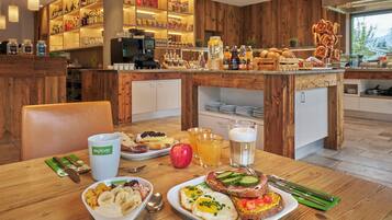 Disposição de mesa de café da manhã com diversos alimentos, máquina de café e área de bar com garrafas e petiscos.