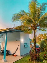 Uma casa pequena, térrea, com fachada branca e detalhes azuis, uma rede e uma palmeira em primeiro plano.