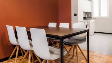 Uma sala de jantar moderna com mesa de madeira e cadeiras brancas, uma luminária pendente e uma cozinha ao fundo.