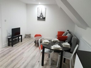 Uma sala moderna com área de jantar, apresentando uma mesa de jantar preta, cadeiras brancas, um sofá com almofadas vermelhas e uma televisão em um suporte preto.