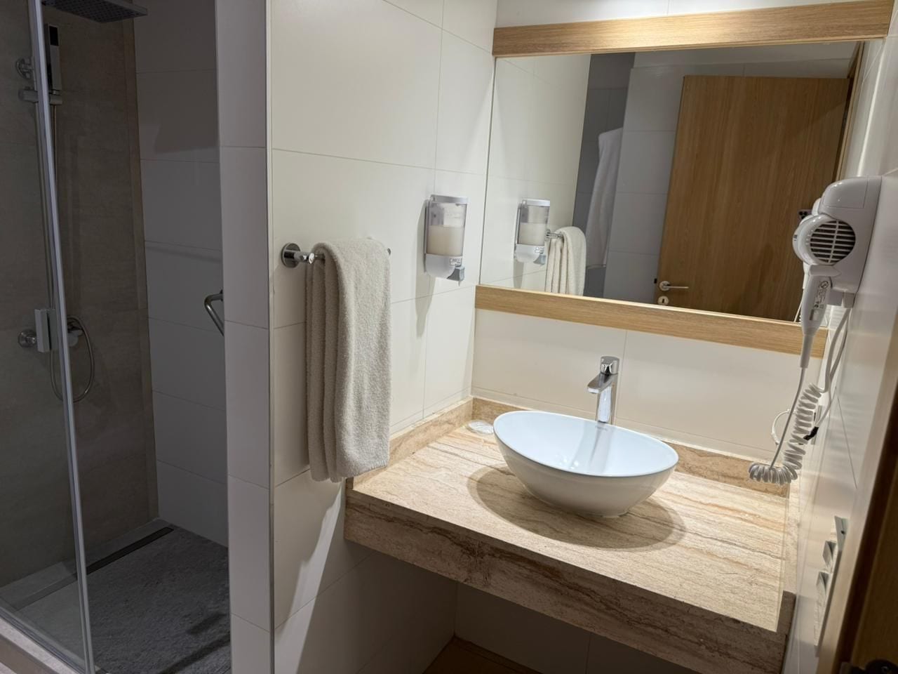 Uma casa de banho com cabine de duche em vidro, toalha bege, lavatório branco com bancada de mármore e secador de cabelo.