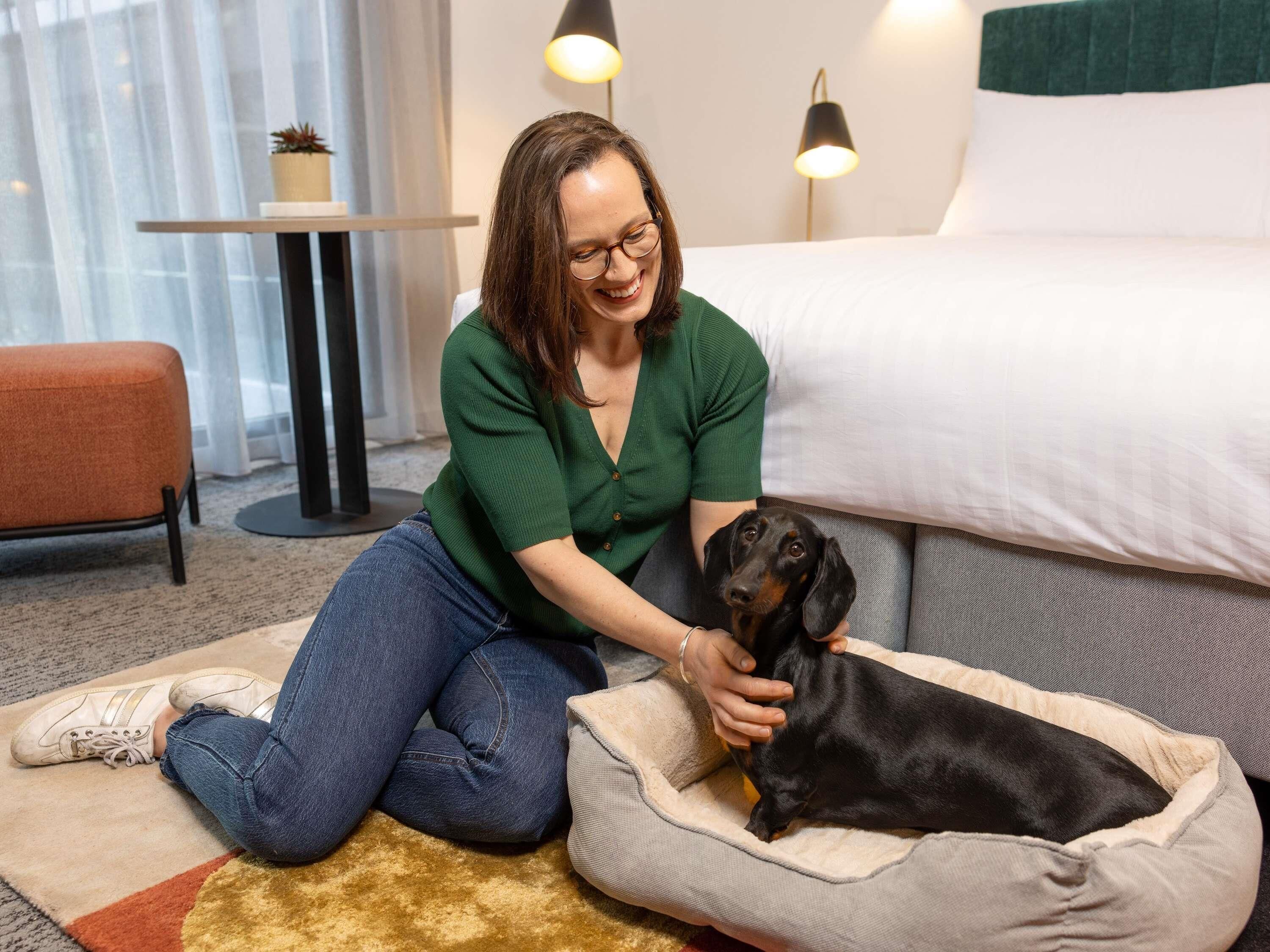 Mulher sentada num tapete ao lado de uma caminha de cachorro em quarto de hotel, com uma cama, uma mesinha e uma luminária.