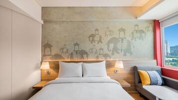 Quarto de hotel moderno com uma cama grande, duas luminárias de cabeceira, um sofá com almofadas e um mural na parede com elefantes e lanternas.