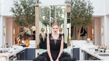 Uma mulher num vestido preto sentada numa sala luxuosa com soalho de mármore, janelas amplas e teto alto com detalhes decorativos.