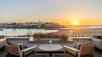 Terraço com mesa e cadeiras com vista para a paisagem urbana costeira ao pôr do sol.