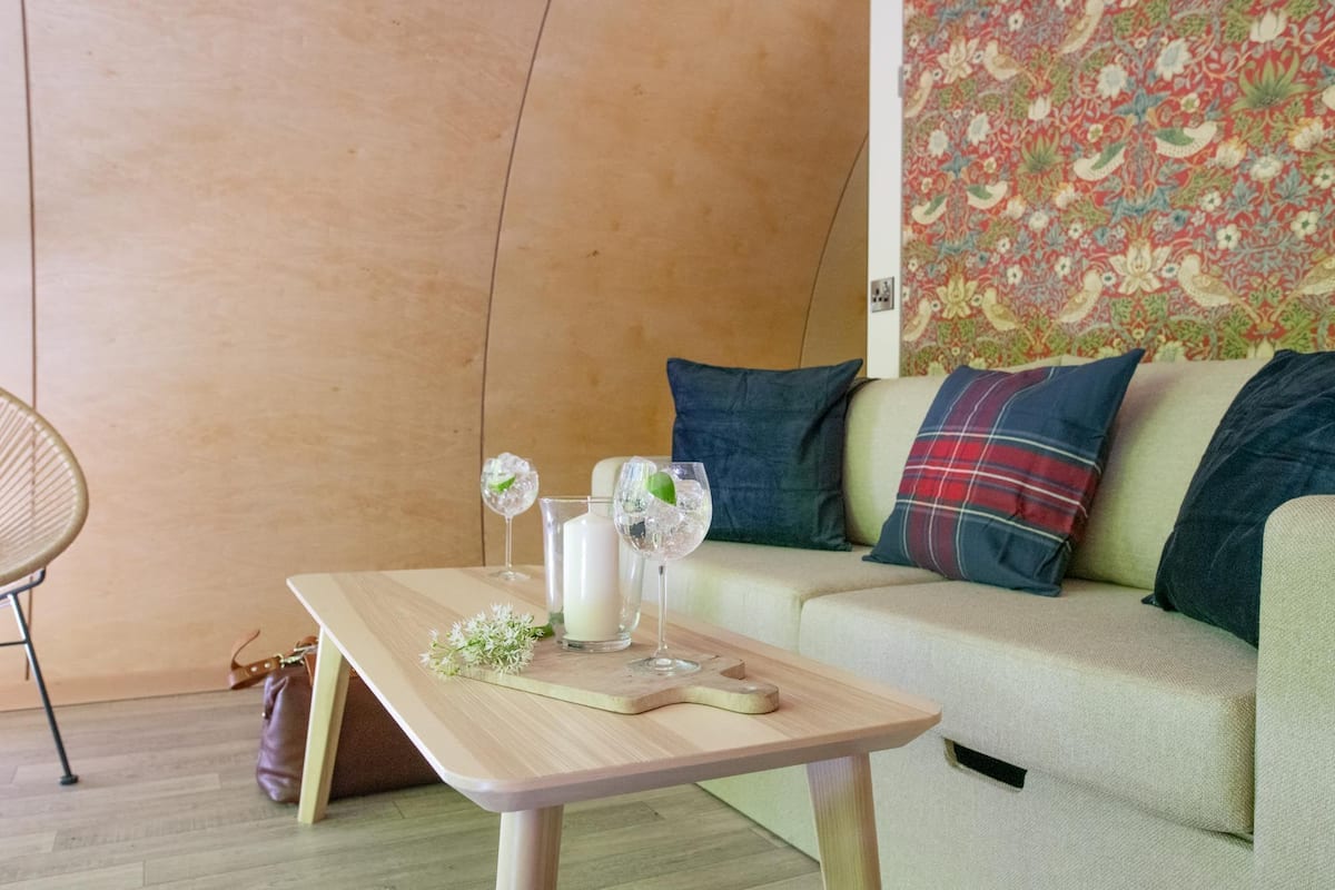 Uma sala de estar moderna com uma mesa de madeira, dois copos com bebida branca e uma planta decorativa. Um sofá com almofadas estampadas está ao fundo.