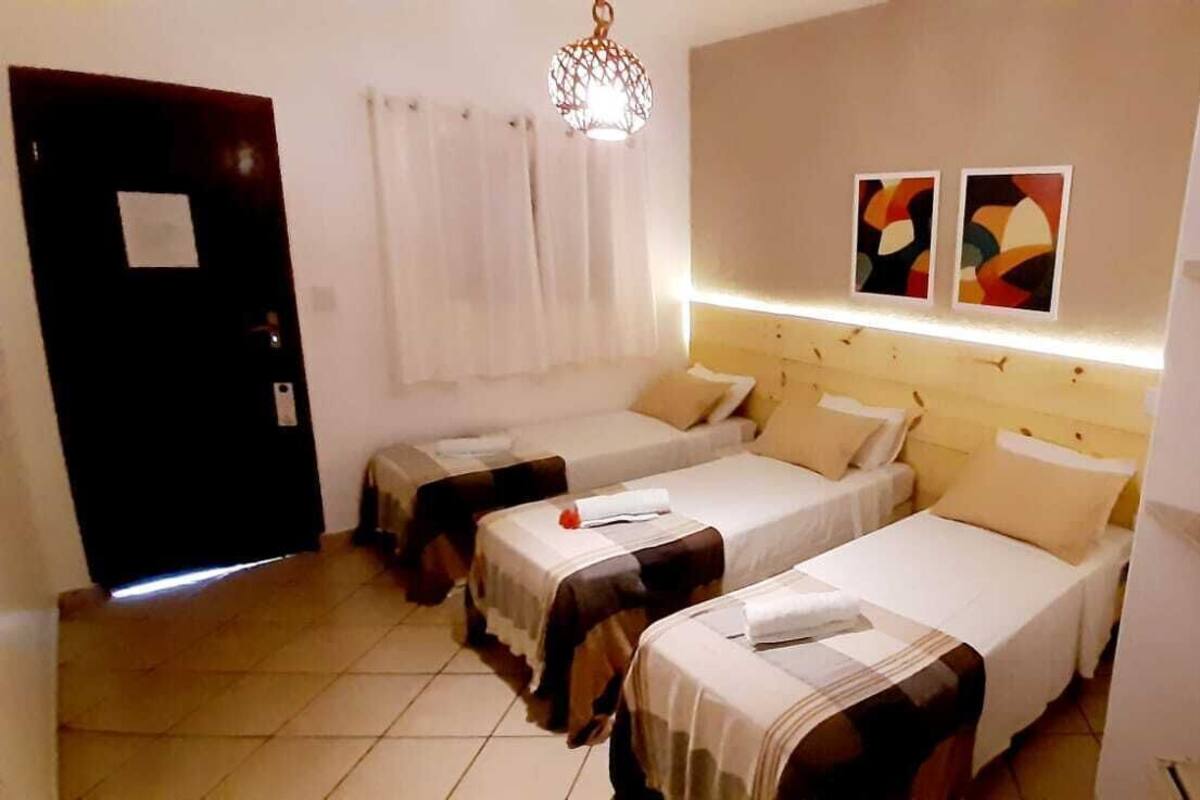 Quarto de hotel com três camas, uma porta, janela com cortinas, luminária fixada na parede e arte abstrata.