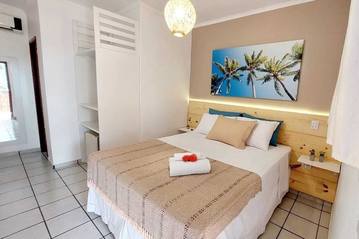 Quarto de hotel com cama, pintura de palmeiras, mesa de cabeceira e criado-mudo.