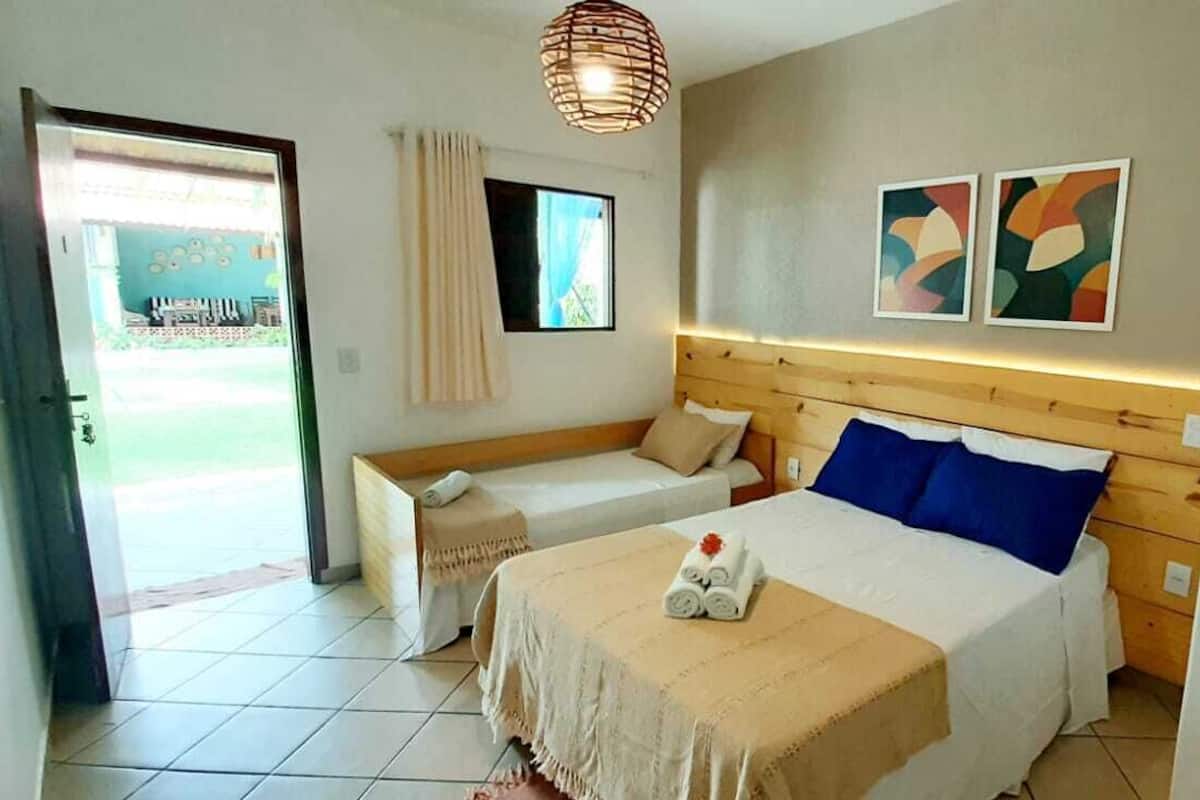 Quarto de hotel com duas camas, cabeceira de madeira, uma janela pequena e porta que dá para uma área externa.