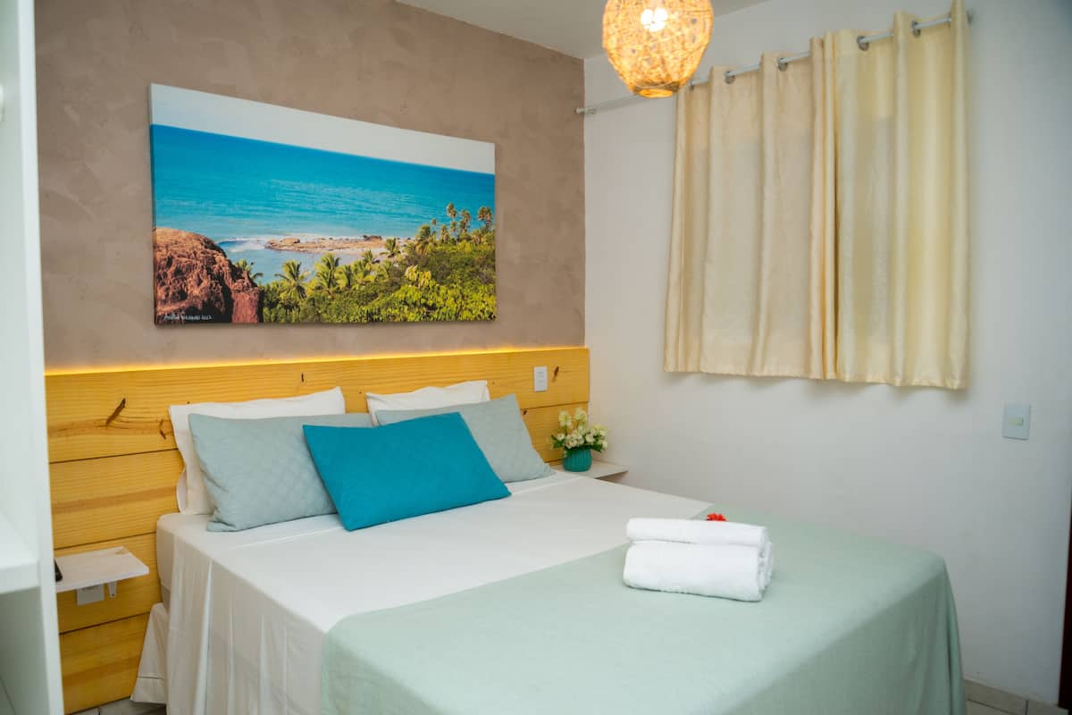 Uma cama bem arrumada com colcha verde, cabeceira de madeira e um quadro com uma paisagem de praia acima.