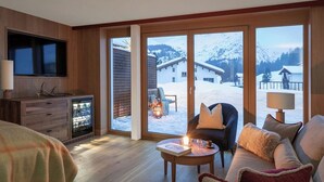 Quarto de hotel com decoração em madeira, televisão, secretária com luminária, sofá e vista para uma paisagem montanhosa coberta de neve através das portas de vidro deslizantes.