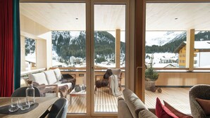 Um interior moderno com um deck de madeira, um sofá, uma mesa de jantar e vista para montanhas nevadas e edifícios.