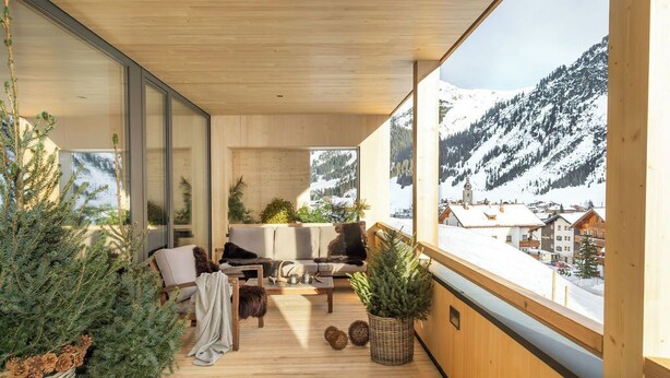 Uma sala de estar moderna com piso de madeira, janelas amplas e vista para uma cidade montanhosa coberta de neve.