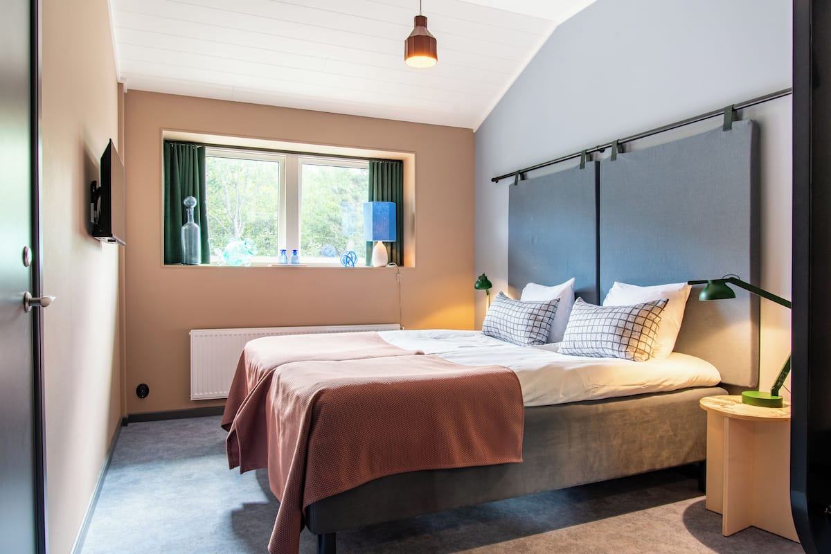 Quarto com cama de casal, mesa de cabeceira, abajur, janela com cortinas e radiador.