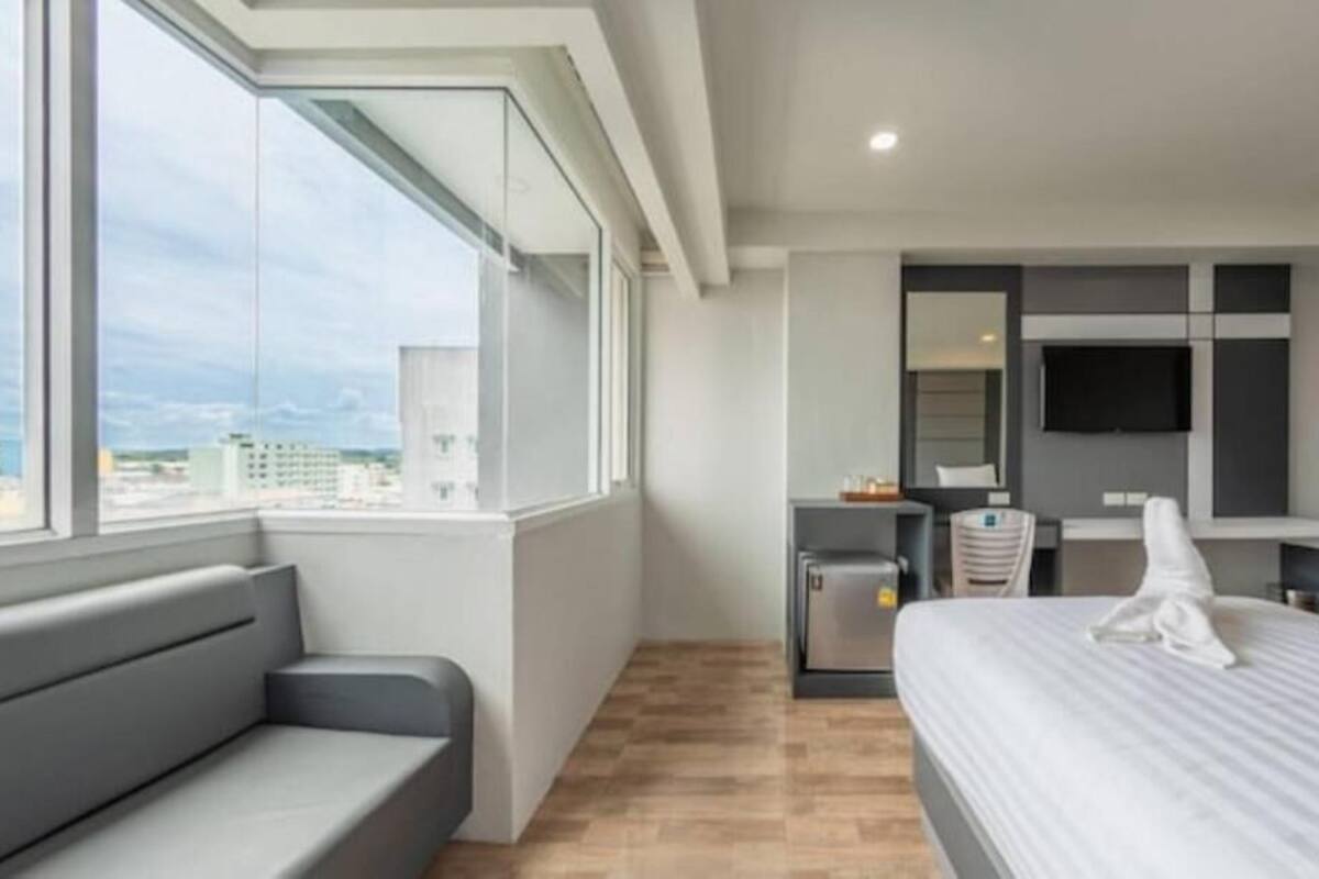 Quarto de hotel moderno com uma janela grande, cama, área de estar, escrivaninha e televisão.