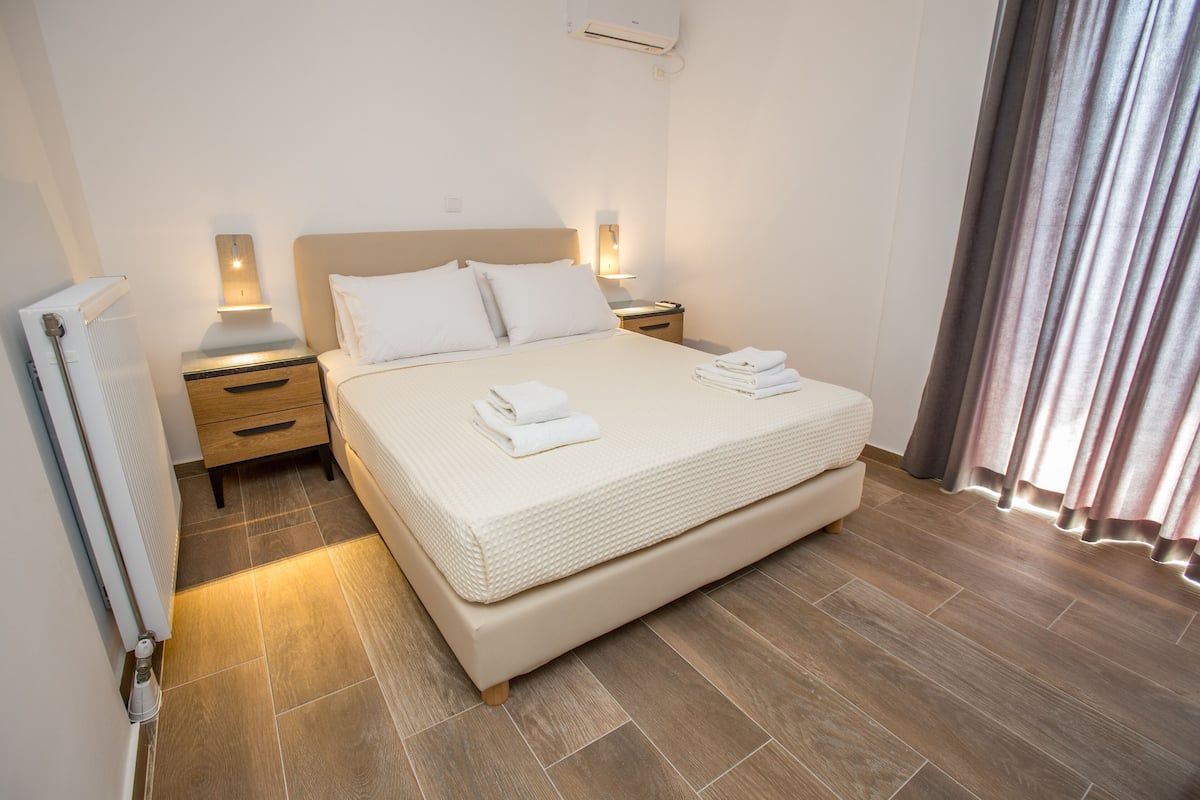 Cama bem arrumada com lençóis brancos, mesa de cabeceira com abajur e radiador à esquerda.