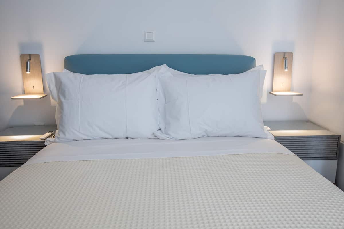Cama bem arrumada com roupa de cama branca e cobertura branca texturizada, com duas mesas de cabeceira e arandelas iluminadas nas paredes.