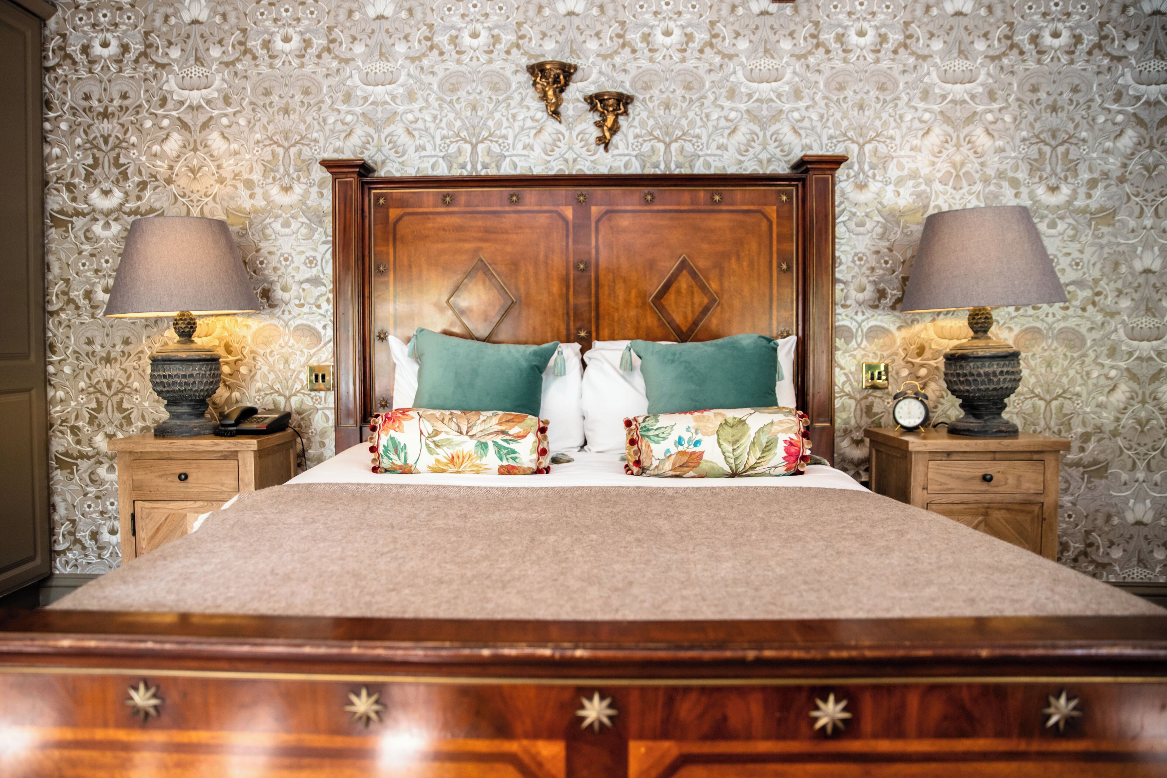 Uma cama com cabeceira de madeira, duas mesas de cabeceira com abajures e um papel de parede estampado com flores.