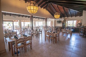 Uma ampla sala de jantar com várias mesas e cadeiras, janelas grandes e teto alto com vigas aparentes.