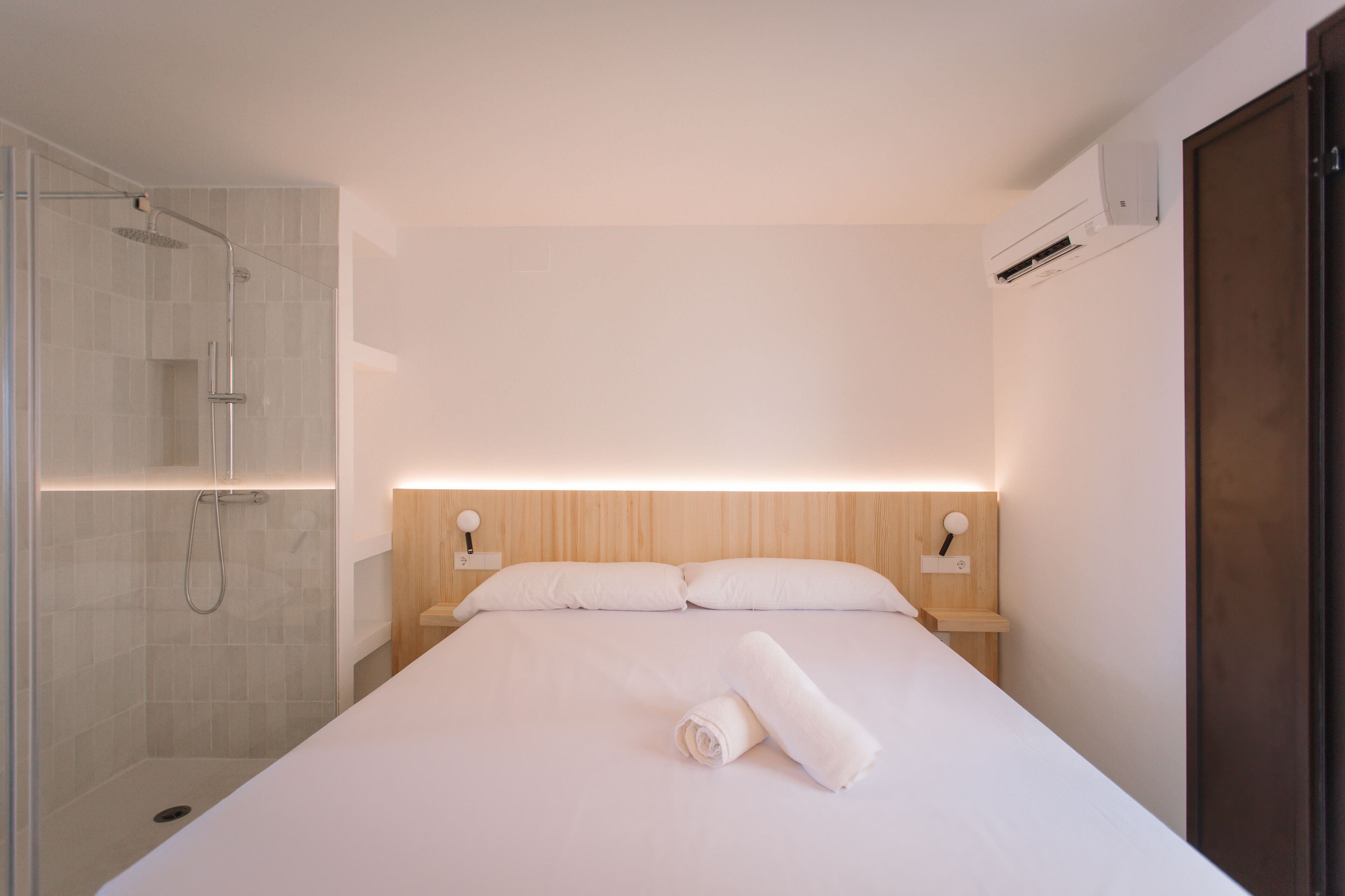 Quarto de hotel moderno com uma cama grande, área de chuveiro e cabeceira de madeira.