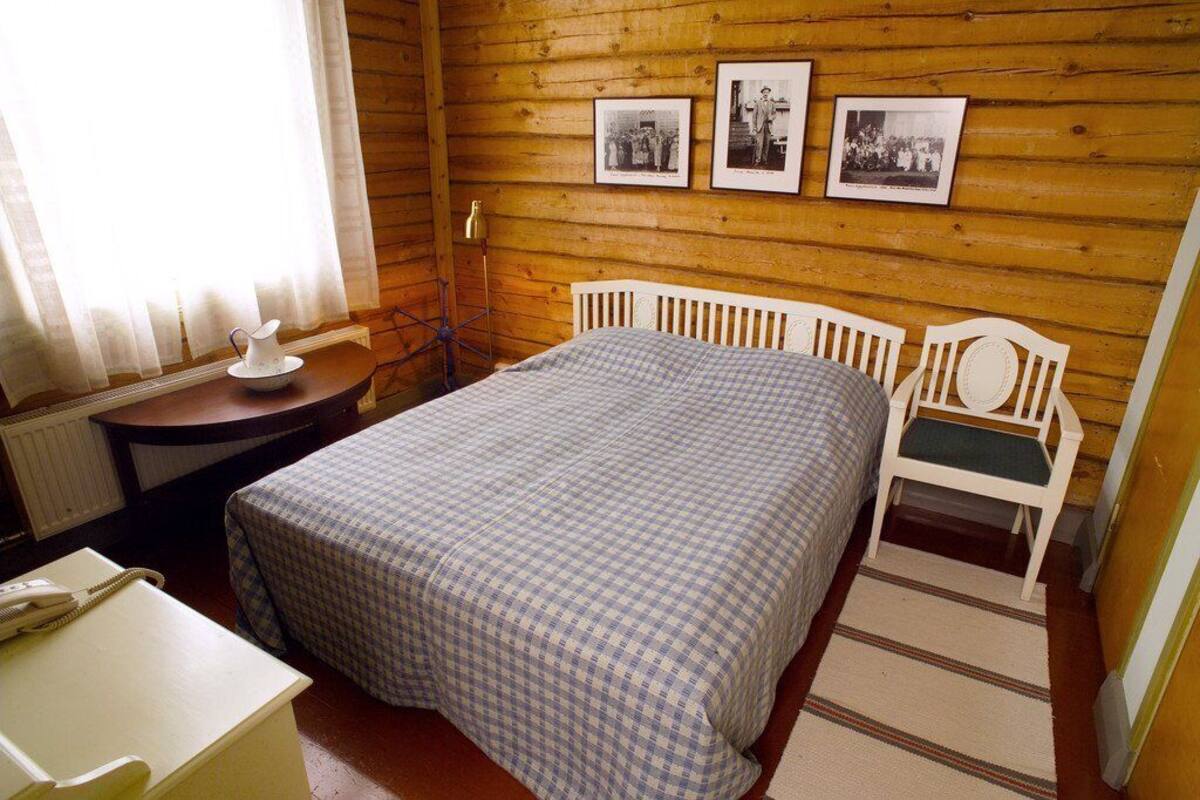 Quarto com colcha xadrez, cabeceira de madeira, mesa de cabeceira com abajur e uma cadeira. Há três quadros na parede.