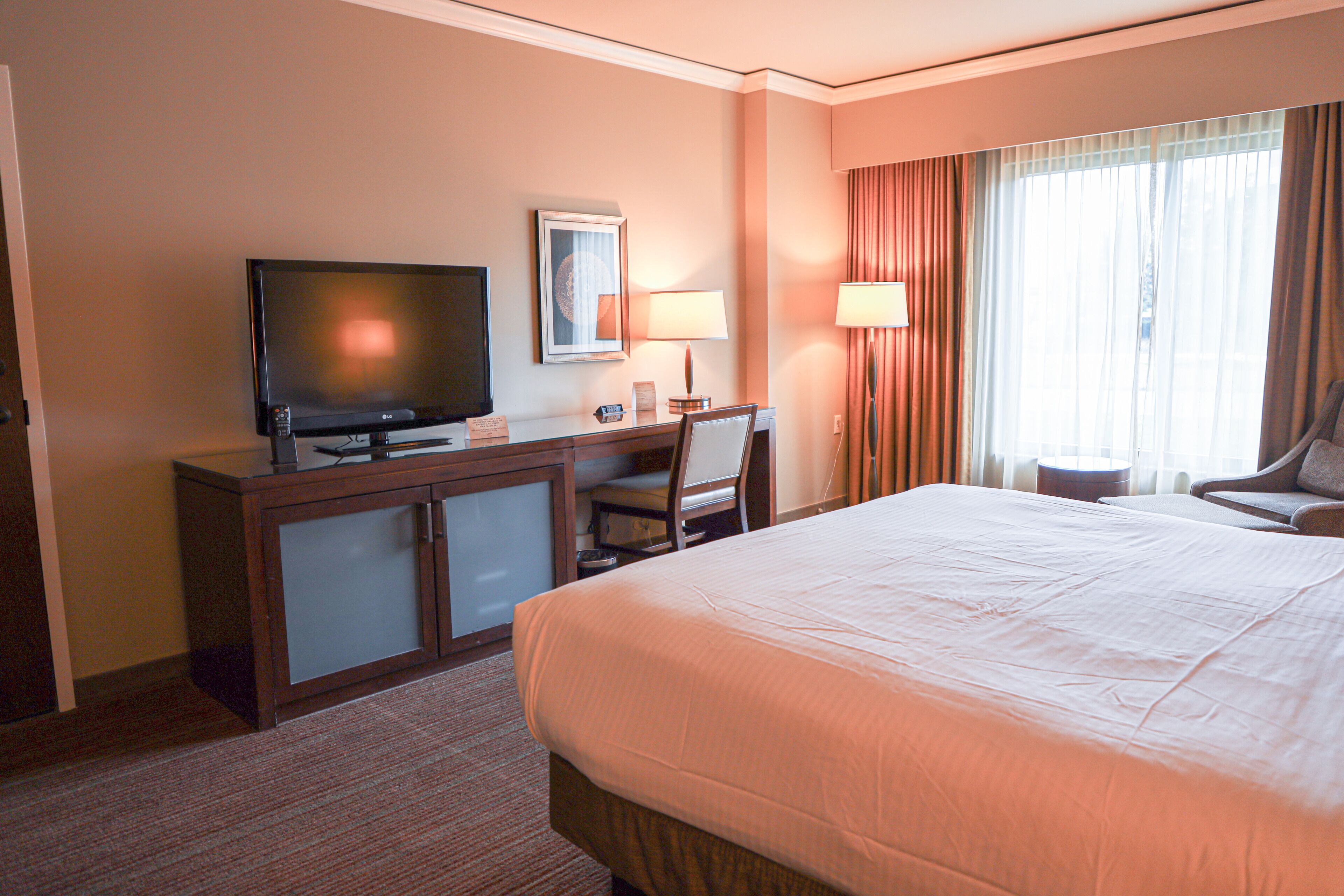 Quarto de hotel com cama, escrivaninha com televisão, cadeira, luminária e janela com cortinas.