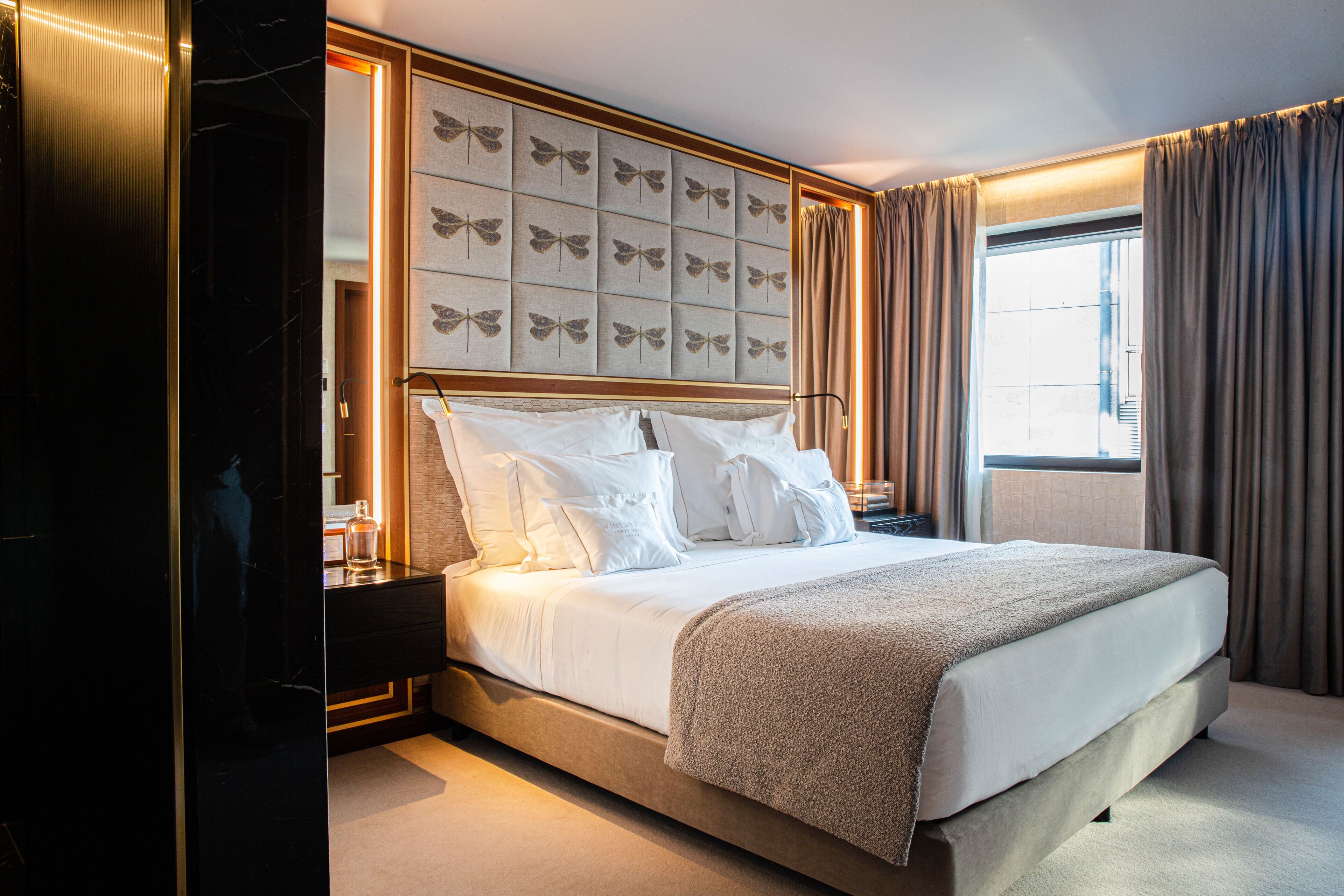 Quarto de hotel com uma cama grande, cabeceira com desenhos de borboletas, mesa de cabeceira e uma janela com cortinas.