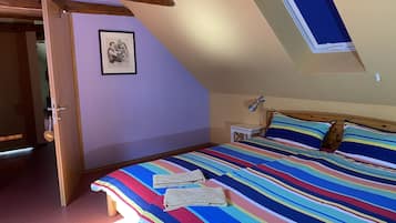 Um quarto com teto inclinado, uma cama com colcha listrada e colorida, duas almofadas, uma mesa de cabeceira com abajur e um quadro na parede.
